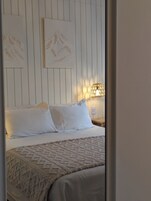 Quarto de hotel com cama, paredes brancas e luminária fixada na parede.