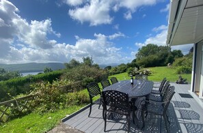 Pátio externo com mesa e cadeiras, com vista para uma paisagem deslumbrante com um lago e montanhas.