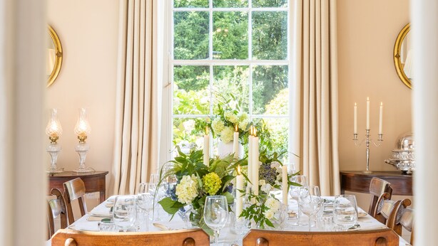 Uma sala de jantar com uma janela grande, um candelabro e uma mesa posta para refeição.