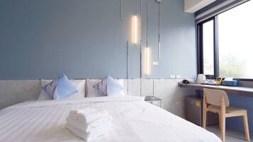 Quarto de hotel moderno com uma cama grande, uma escrivaninha e uma cadeira. Há duas luminárias fixadas na parede e uma janela com vista para árvores.
