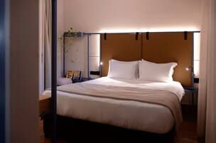 Cama bem arrumada com lençóis brancos, mesa de cabeceira com abajur e prateleira na parede com uma planta.