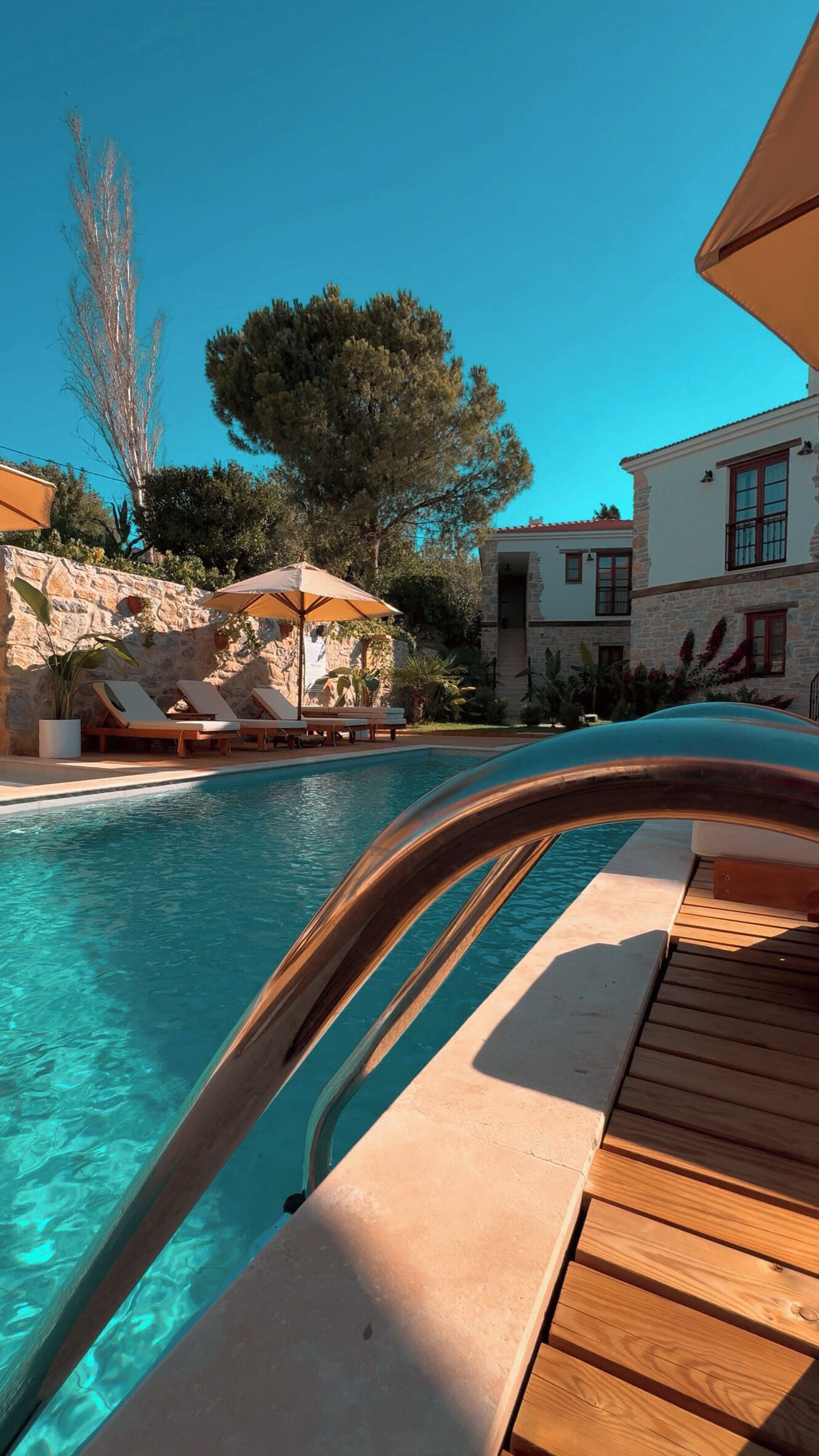 Piscina com deck de madeira, espreguiçadeiras e muro de pedra.