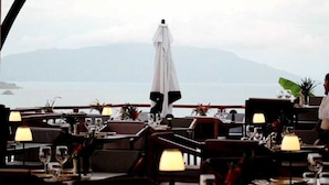 Restaurante com mesas preparadas para refeições, um garçom e vista para o mar e montanhas ao fundo.