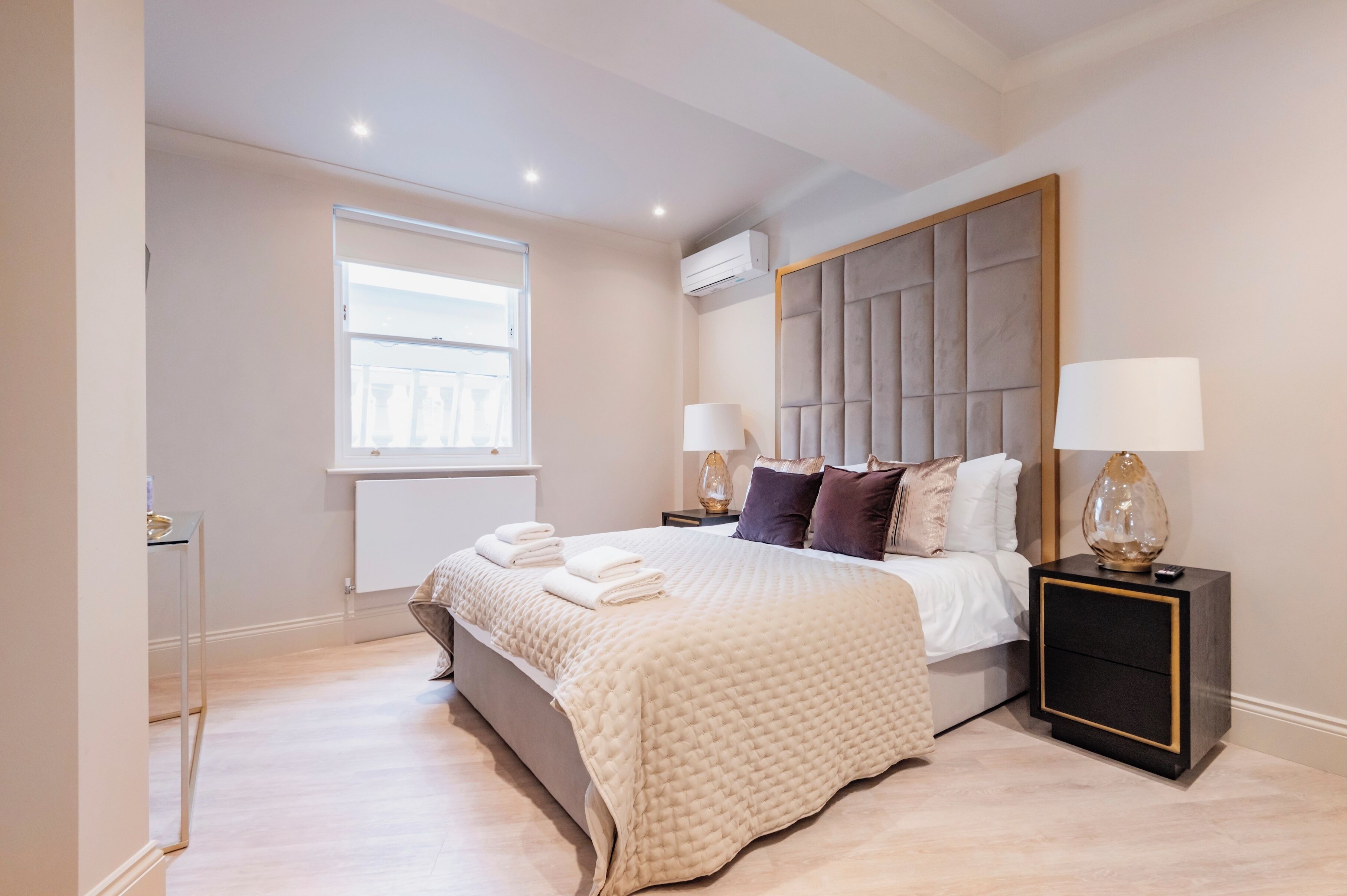 Um quarto moderno com uma cama grande, duas mesas de cabeceira com luminárias, uma janela com persianas e um aparelho de ar condicionado na parede.