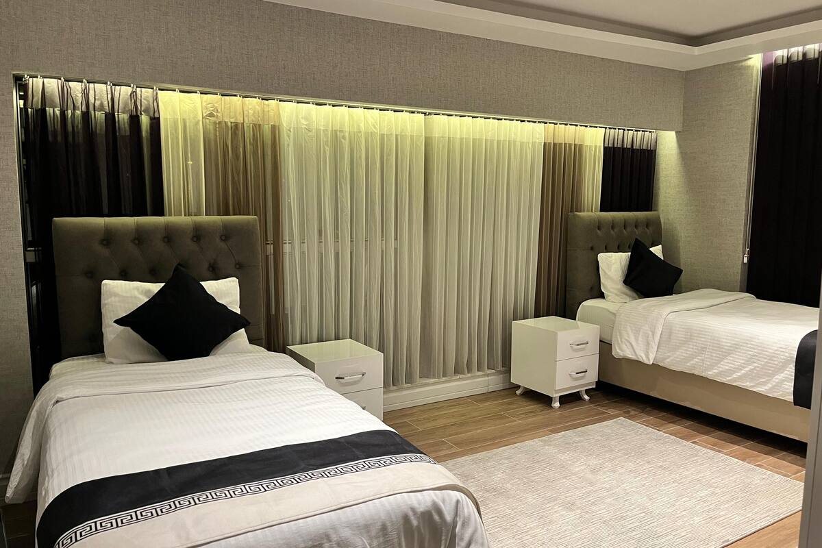 Quarto de hotel com duas camas, uma mesinha de cabeceira e uma janela grande com cortinas.