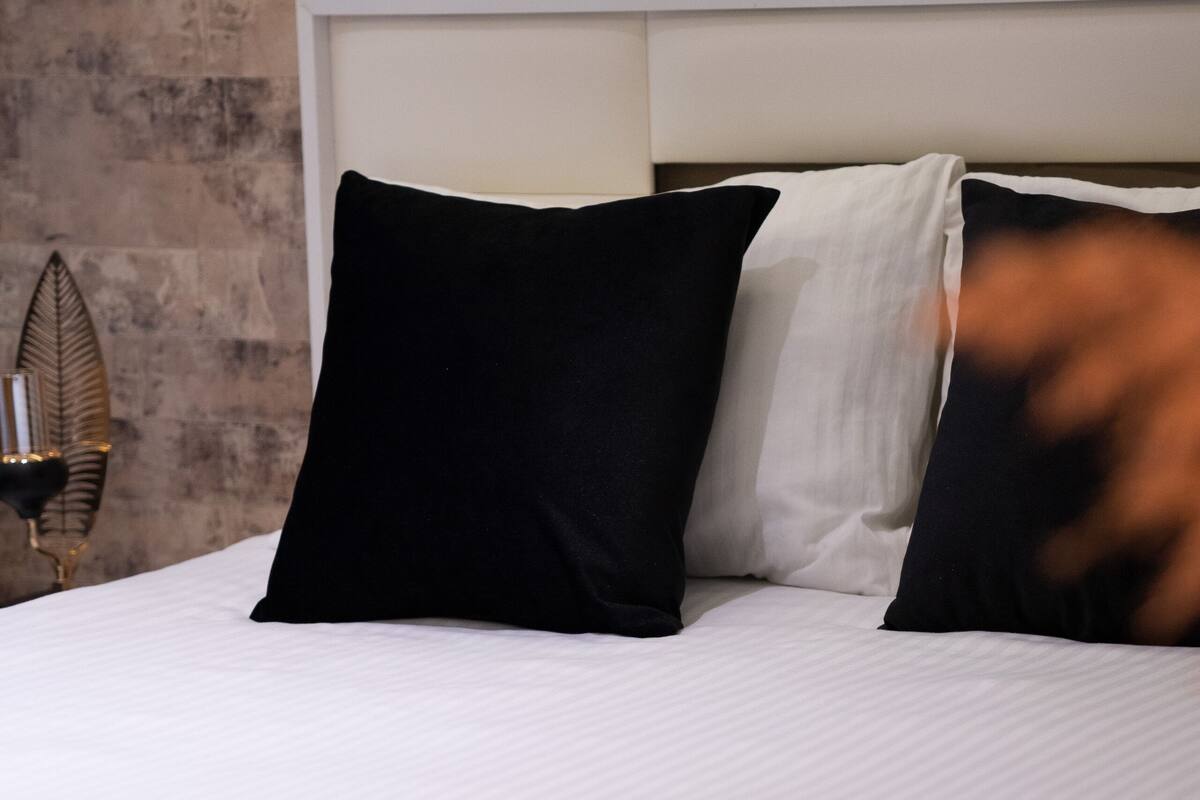 Cama com roupa de cama branca e almofadas preto e branco, diante de uma parede texturizada.