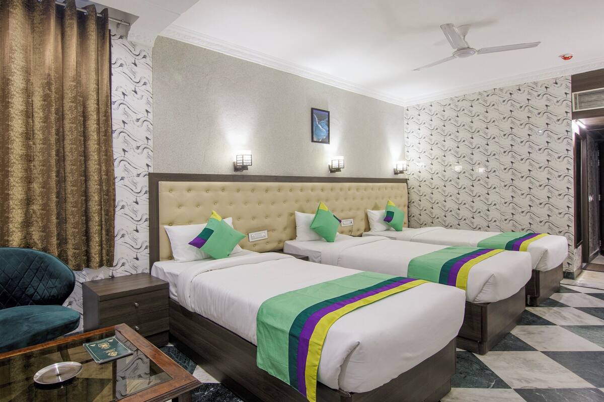 Quarto de hotel com duas camas, uma poltrona verde, uma mesinha de centro com um livro e um ventilador de teto.