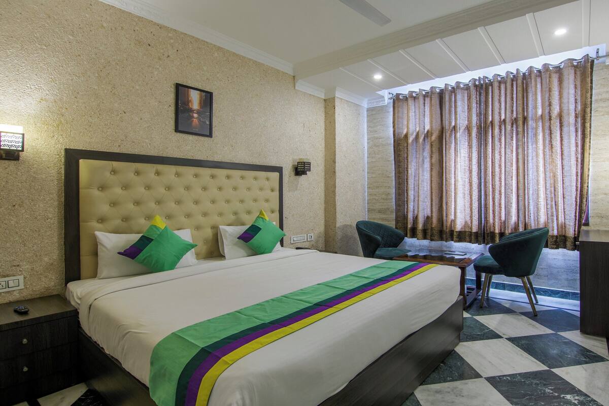 Quarto de hotel com uma cama grande, uma escrivaninha, duas cadeiras, uma janela com cortinas e um quadro na parede.