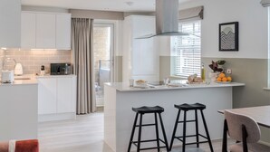 Uma cozinha moderna com armários brancos, uma ilha central com banquetas e uma janela grande com cortinas.