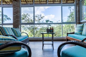 Quarto com mobiliário de vime, mesa de vidro com garrafas e vista para árvores através de grandes janelas.