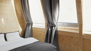 Quarto com teto de madeira, cama com roupa de cama branca e cinza e janela com cortinas cinzas.