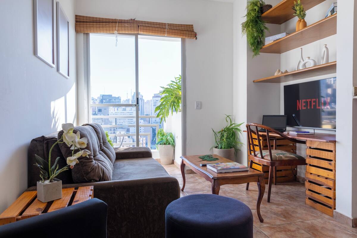 Sala de estar moderna com sofá, mesa de centro, televisão e vista para a cidade através de uma grande janela.