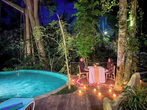Uma área de refeições ao ar livre com piscina, rodeada por vegetação tropical e iluminada por velas.