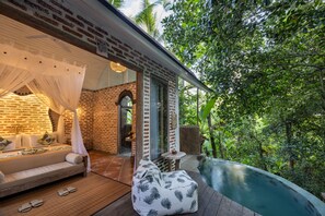 Uma área de estar externa aconchegante com sofá e almofadas, uma mesa pequena e um banco, tudo em um deck de madeira com vista para um jardim exuberante e uma piscina.