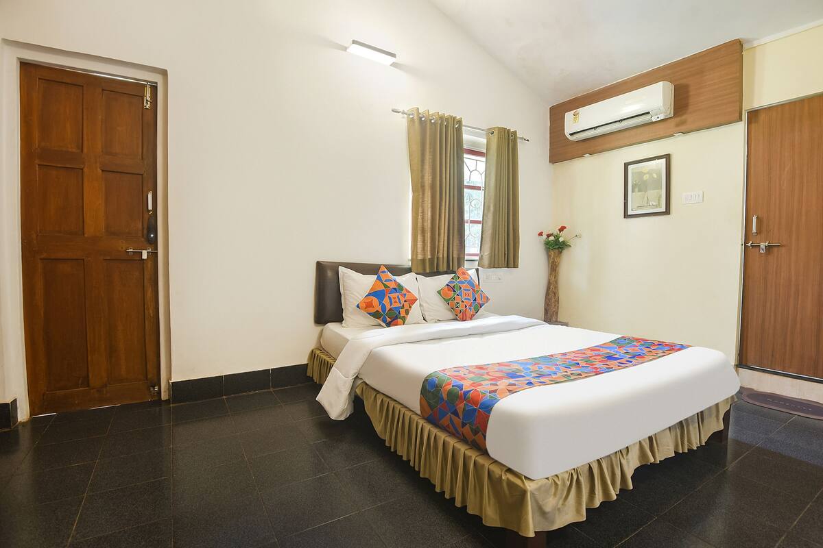 Quarto de hotel com uma cama grande, porta de madeira, janela com cortinas, unidade de ar condicionado e uma pequena planta.