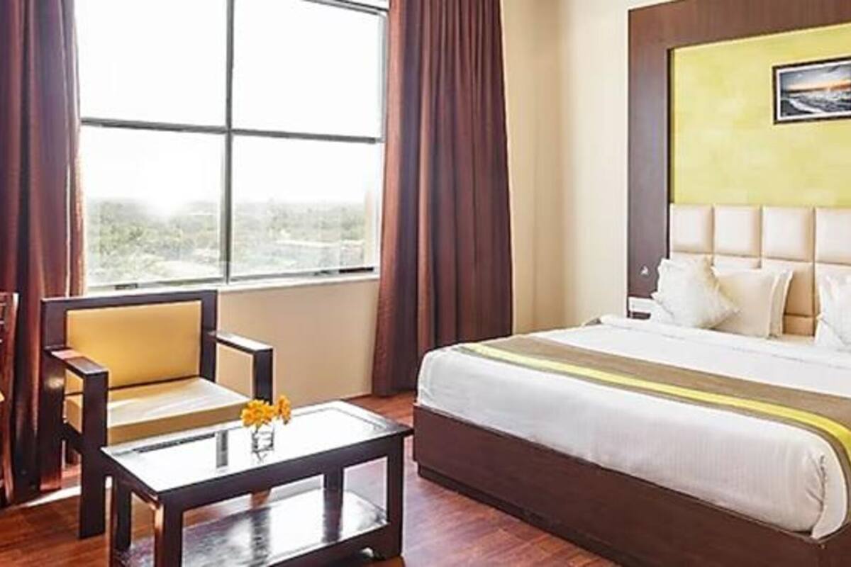 Quarto de hotel com uma cama grande, uma mesa de cabeceira de madeira, uma pequena mesa com um vaso de flores e vista para o exterior pela janela.