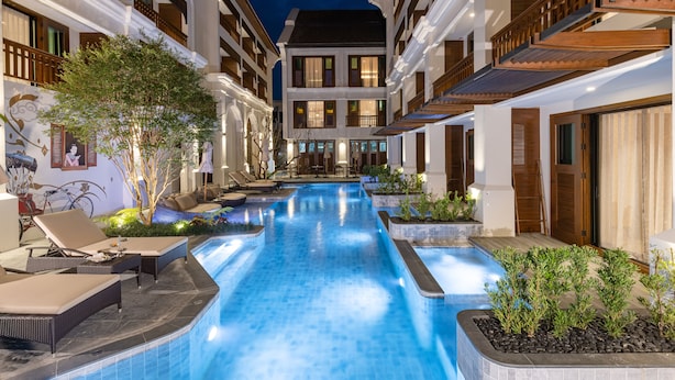 Uma piscina com espreguiçadeiras e plantas, cercada por edifícios com várias andares e varandas.