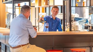 Uma área de bar com um bartender servindo uma bebida, prateleiras com garrafas de bebidas alcoólicas e uma placa indicando uma licença.