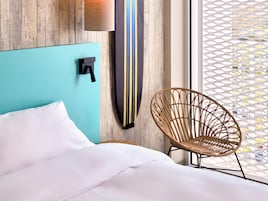 Uma cama com roupa de cama branca, uma luminária fixada na parede e uma cadeira de vime.