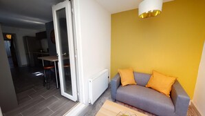 Uma sala de estar moderna com um sofá cinza, duas almofadas mostarda e uma mesa de centro de madeira. Um radiador branco está encostado na parede, e uma porta de vidro dá acesso a uma área de cozinha com mesa e banquetas.