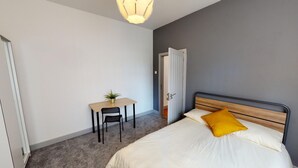 Quarto com cama, escrivaninha, cadeira, uma plantinha e porta.
