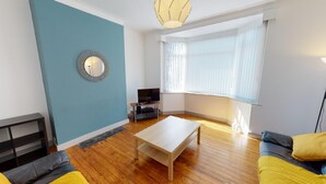 Uma sala de estar moderna com piso de madeira, uma TV sobre um suporte, um espelho redondo e uma janela grande com persianas.