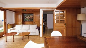 Uma sala de estar ampla com um sofá branco, duas mesas de centro de madeira e uma mesa de jantar de madeira. Uma porta de madeira dá acesso a uma cozinha, e um quadro está visível na parede.