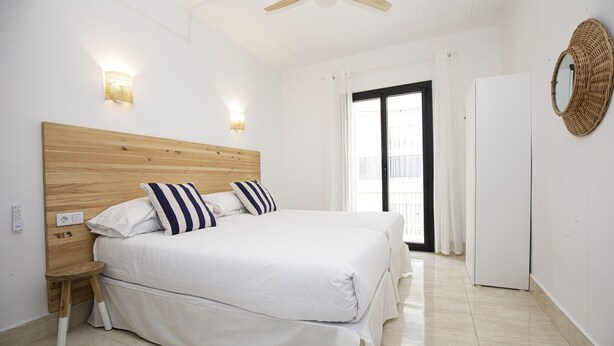 Quarto com cabeceira de madeira, cama com roupa de cama branca, um pequeno banquinho de madeira, guarda-roupa branco e um ventilador de teto.