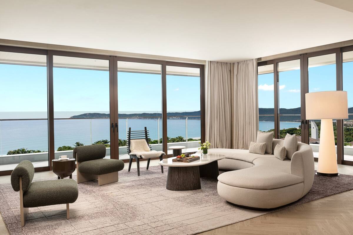Sala de estar moderna com uma janela ampla que oferece vista para o mar e colinas. Há duas poltronas, um sofá e uma mesa de centro.