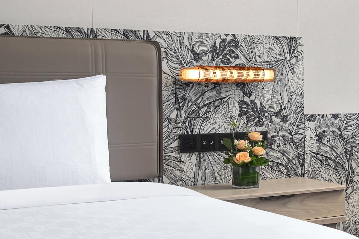 Cama bem arrumada com travesseiro branco e cabeceira, em frente a uma parede com papel de parede estampado com folhagens. Um criado-mudo com um vaso de flores e uma luminária também são visíveis.