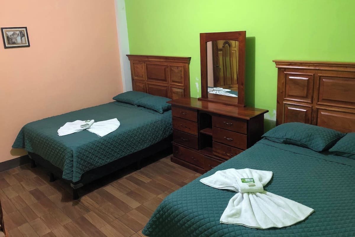 Quarto de hotel com duas camas de solteiro, cada uma com um edredom verde e uma toalha branca. Há uma cômoda de madeira com gavetas e um espelho em cima. As paredes são em tons de pêssego e verde, e o chão é de madeira.