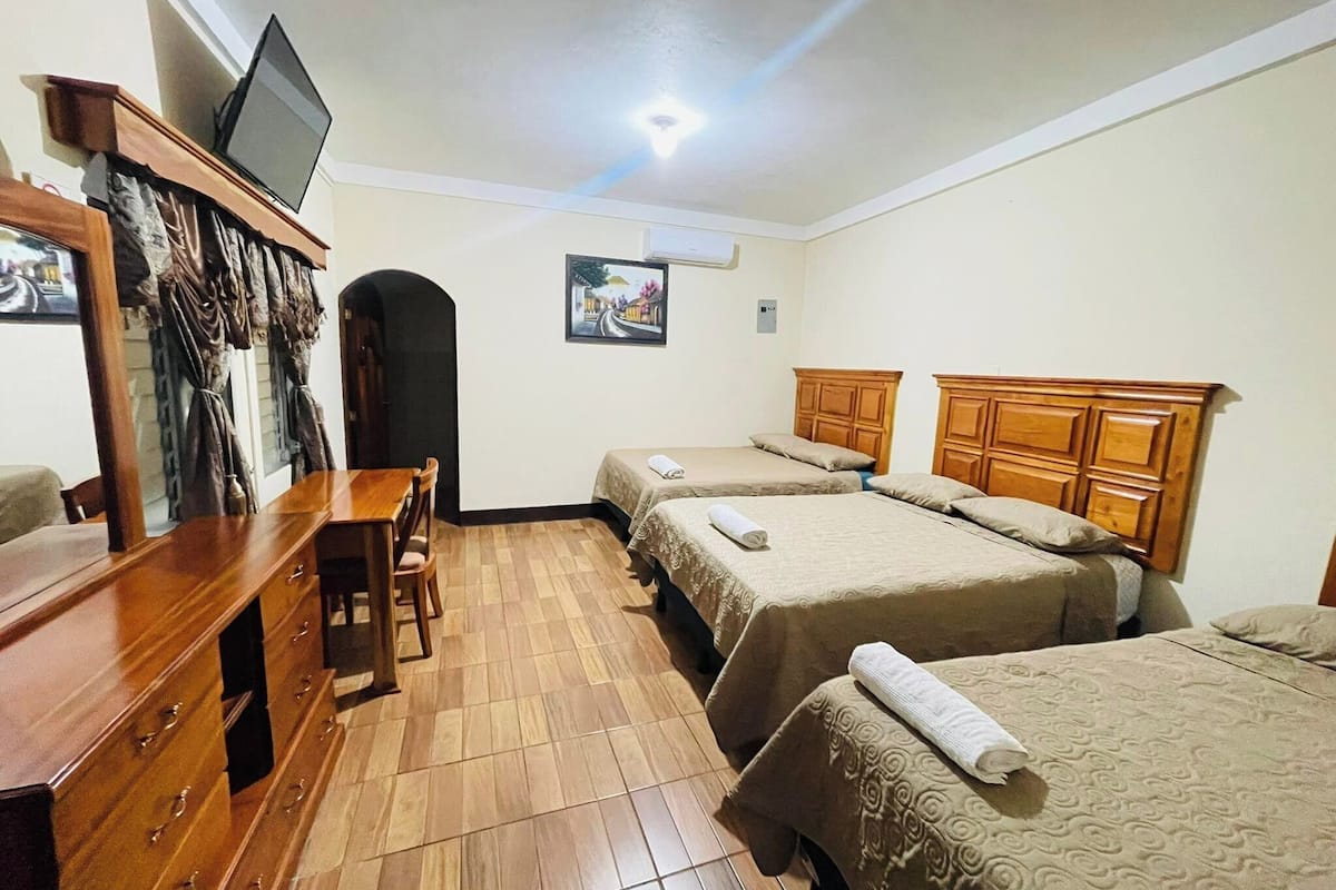 Quarto de hotel com duas camas, cômoda de madeira, escrivaninha, cadeira, televisão na parede e um quadro na parede.