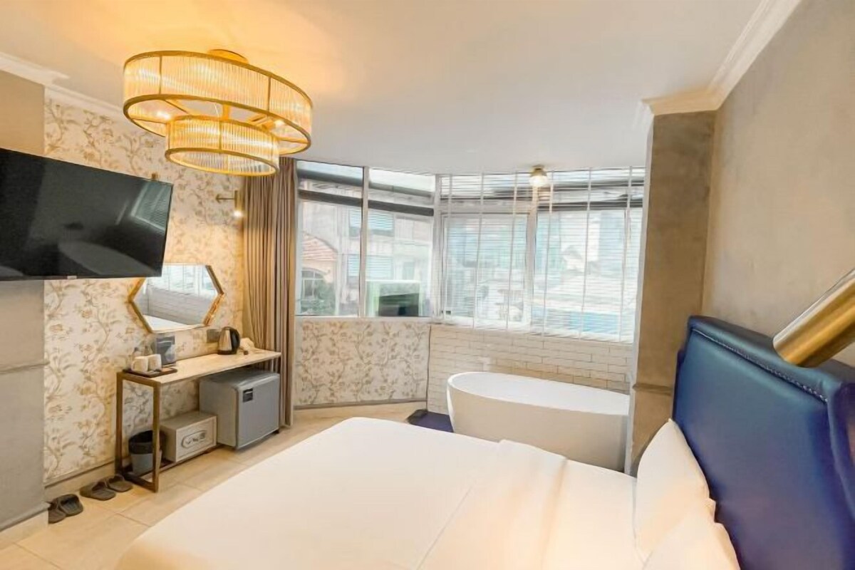 Quarto de hotel com cama, escrivaninha, televisão, janela e banheira.