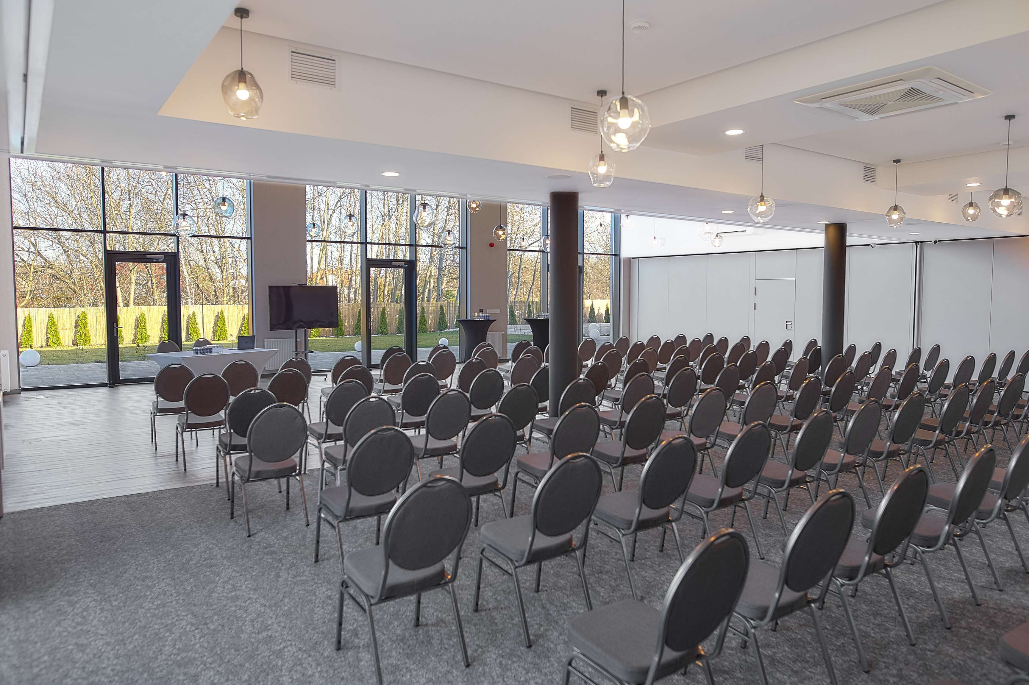 Uma sala de conferências com fileiras de cadeiras, uma tela grande e vista para árvores através de janelas amplas.