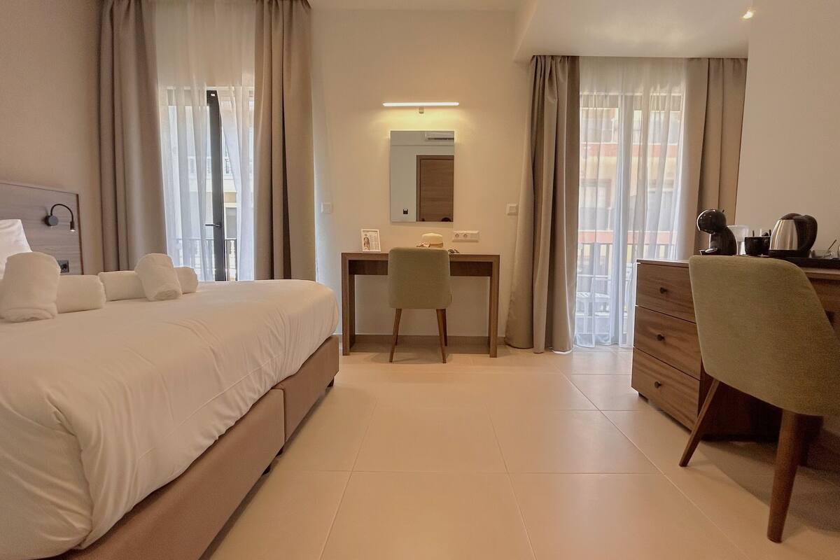 Quarto de hotel com cama, escrivaninha, cadeira, cômoda e varanda com cortinas.