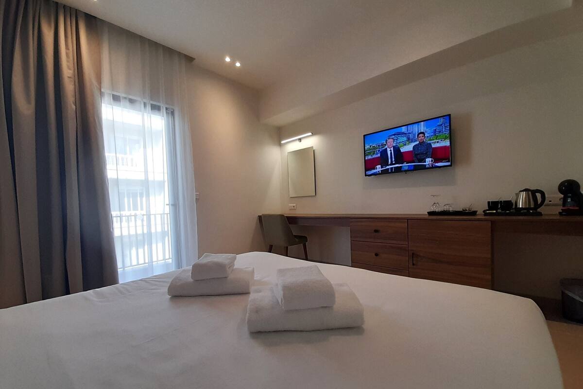 Quarto de hotel com cama, televisão na parede, secretária com cadeira e janela com cortinas.