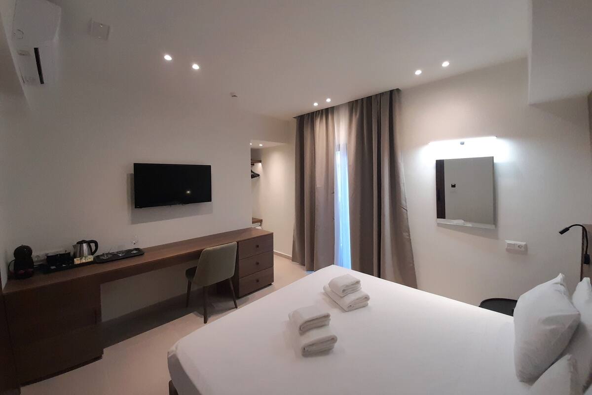 Quarto de hotel com cama, escrivaninha, cadeira, televisão e espelho.