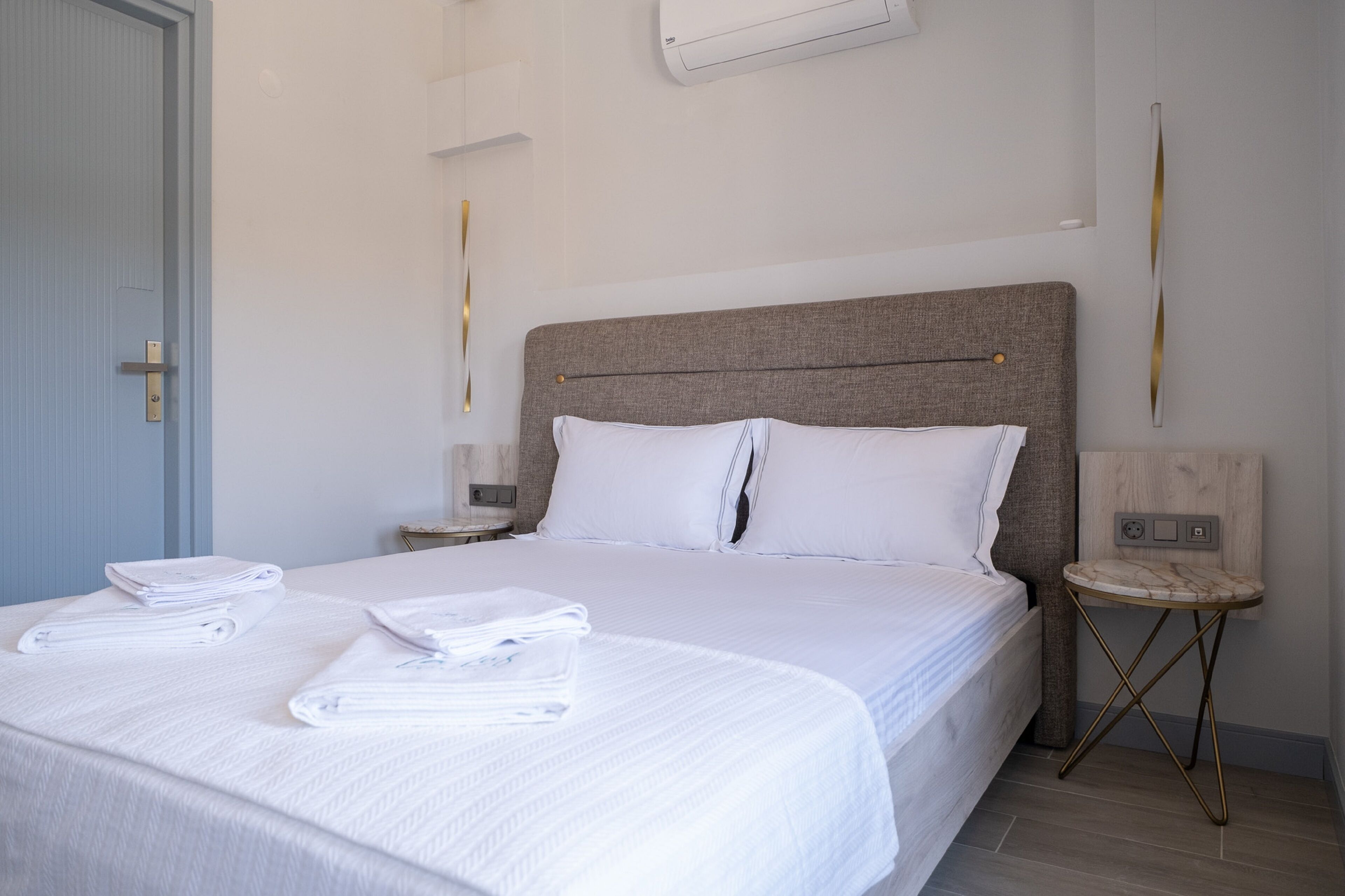 Uma cama bem arrumada com lençóis e toalhas brancas, uma mesa de cabeceira com abajur e um ar condicionado fixado na parede.