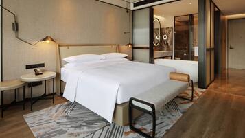 Quarto de hotel moderno com uma cama grande, mesa de cabeceira com abajur, uma pequena mesa redonda e banheiro visível através de uma porta de vidro.
