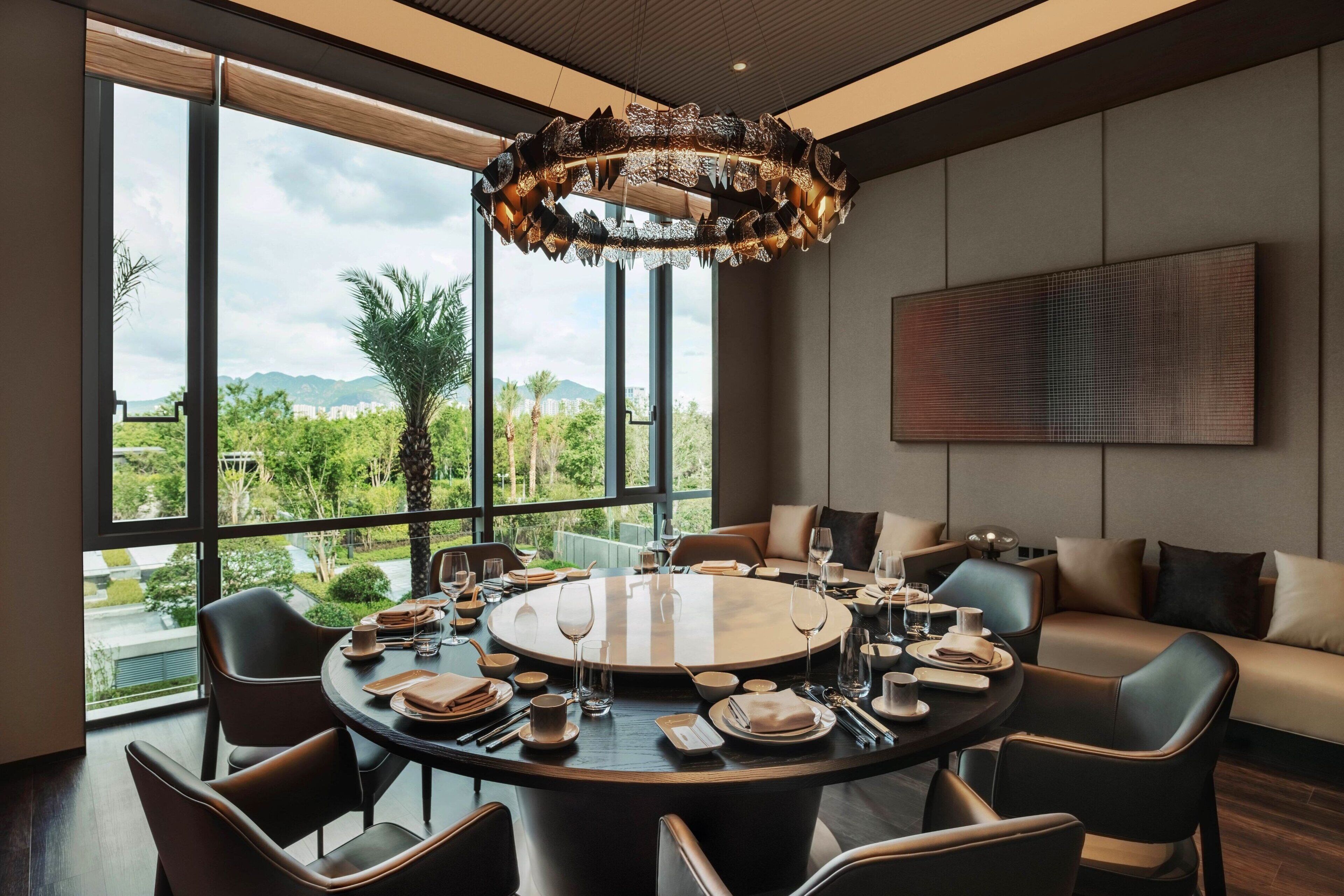 Uma sala de jantar moderna com uma mesa redonda posta para seis pessoas, grandes janelas com vista para vegetação e montanhas, e um lustre contemporâneo.