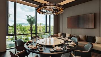 Uma sala de jantar moderna com uma mesa redonda posta para seis pessoas, grandes janelas com vista para vegetação e montanhas, e um lustre contemporâneo.