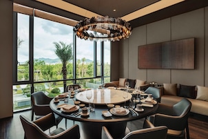 Uma sala de jantar moderna com uma mesa redonda posta para seis pessoas, grandes janelas com vista para vegetação e montanhas, e um lustre contemporâneo.