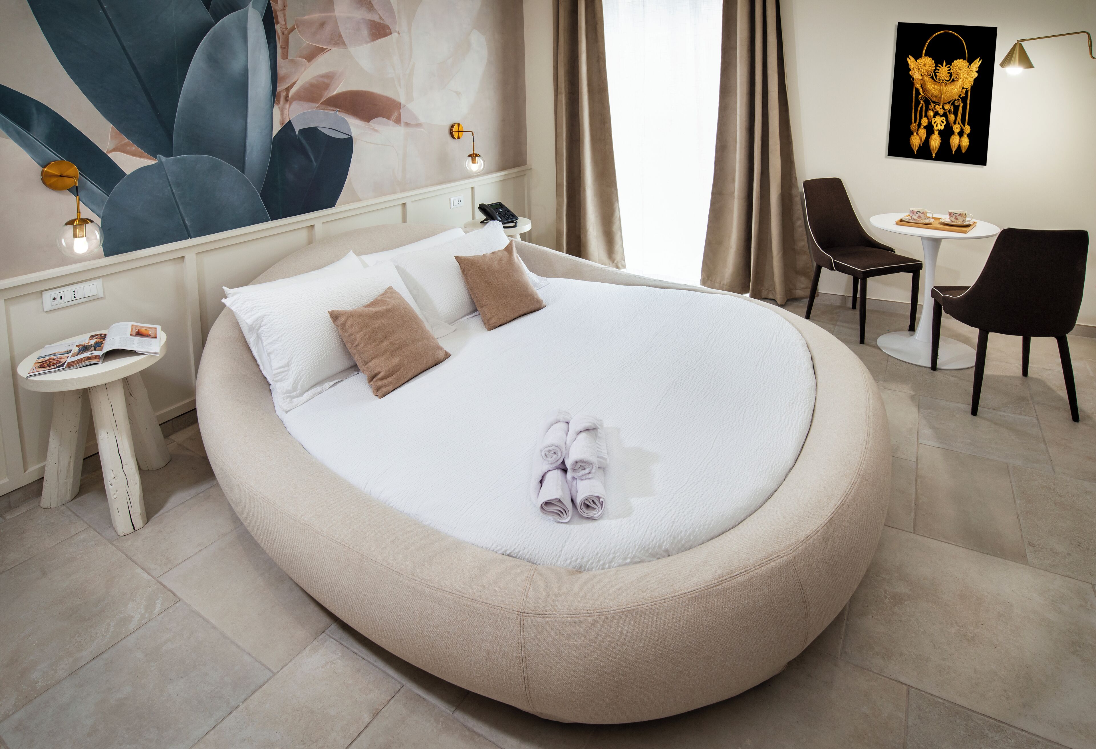 Quarto de hotel moderno com uma cama circular única, uma pequena mesa redonda, duas cadeiras, uma luminária de parede e um grande quadro abstrato.