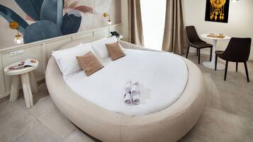 Quarto de hotel moderno com uma cama circular única, uma pequena mesa redonda, duas cadeiras, uma luminária de parede e um grande quadro abstrato.
