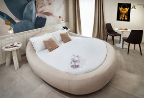 Quarto de hotel moderno com uma cama circular única, uma pequena mesa redonda, duas cadeiras, uma luminária de parede e um grande quadro abstrato.