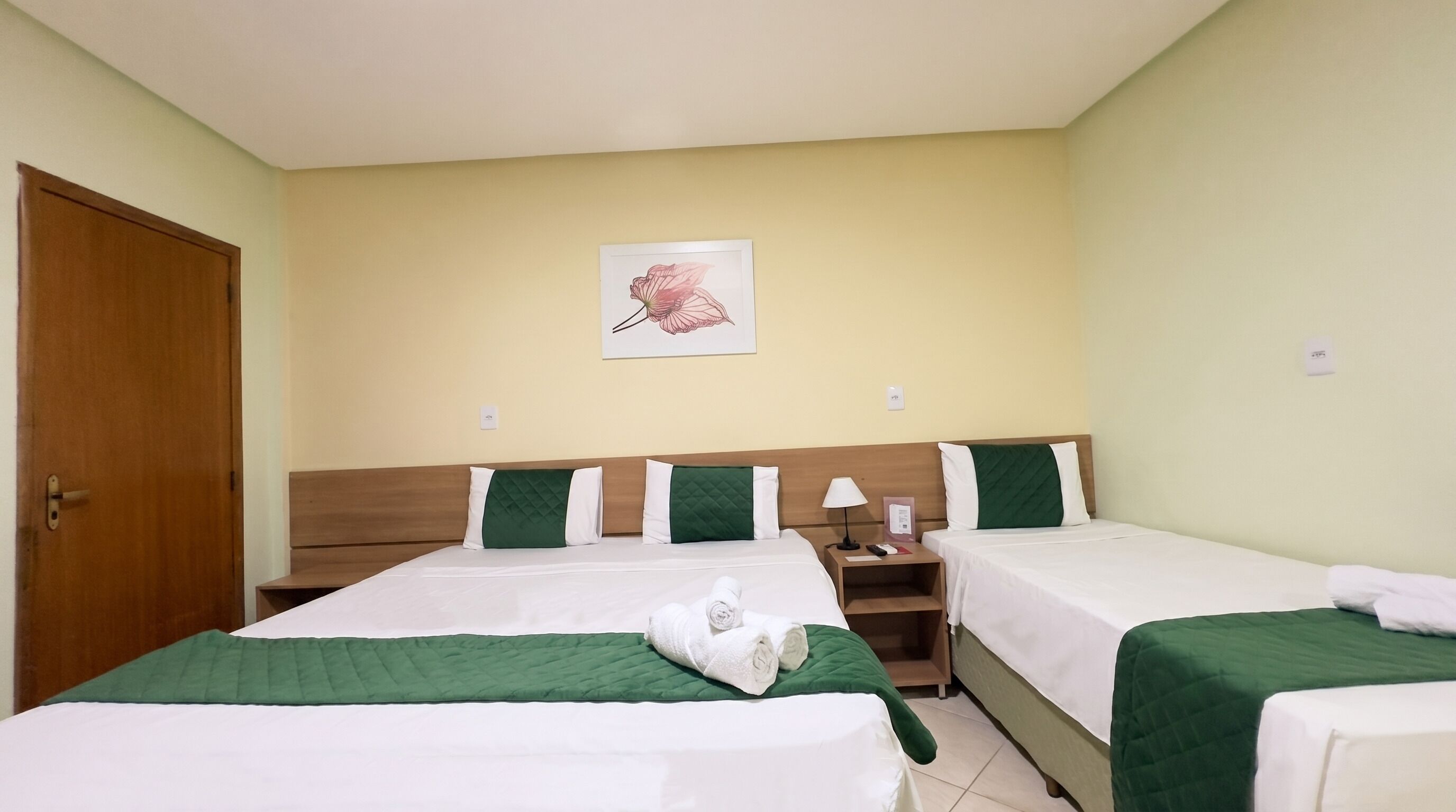 Quarto de hotel com duas camas, cabeceira de madeira, mesa de cabeceira, luminária e quadro pendurado na parede.