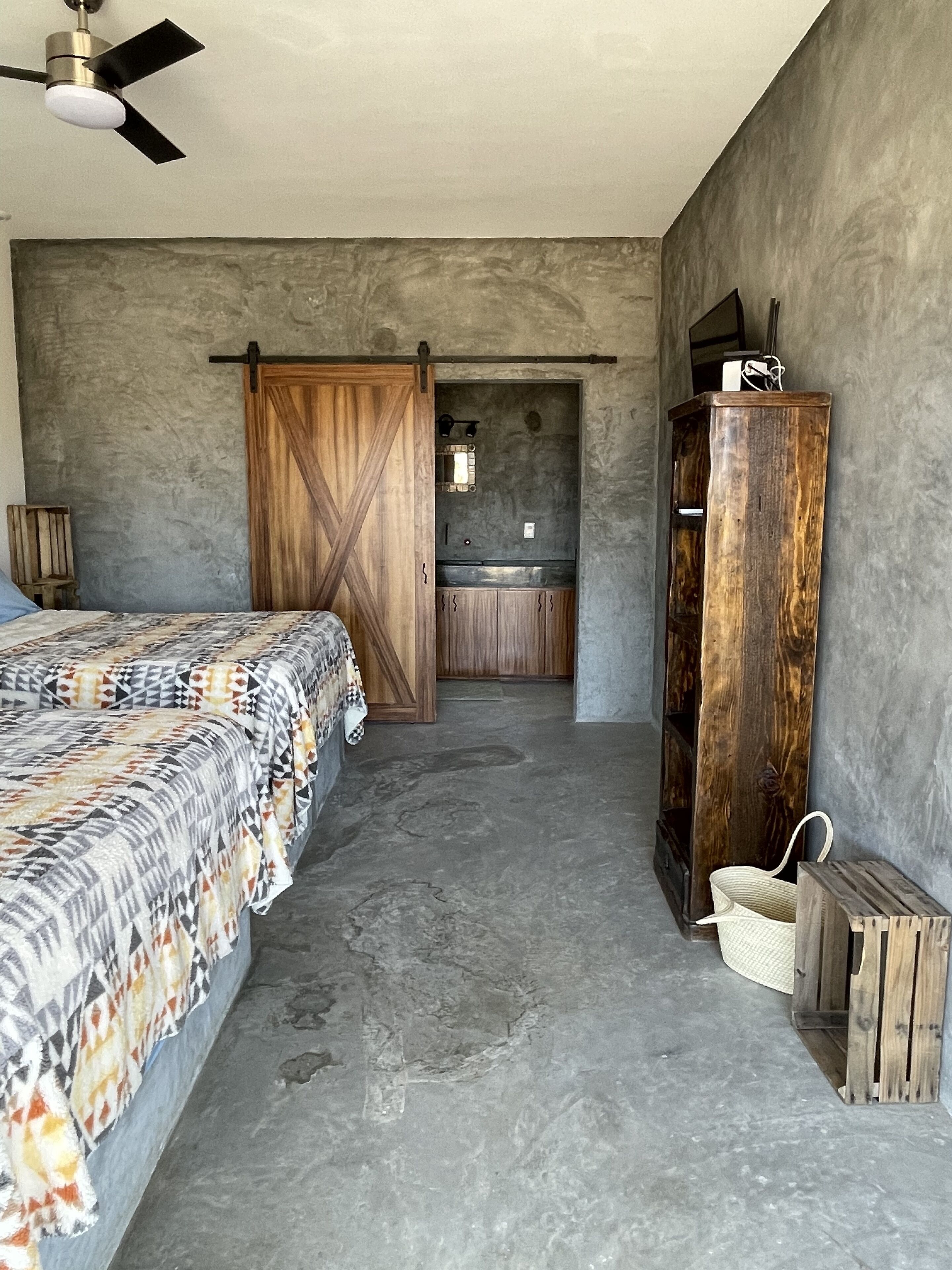Quarto com cama, guarda-roupa de madeira, pequena cozinha e ventoinha no teto.
