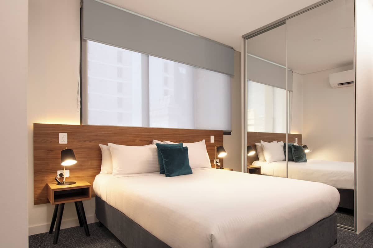 Quarto de hotel com uma cama grande, duas mesas de cabeceira com luminárias, um espelho e uma janela com persianas.