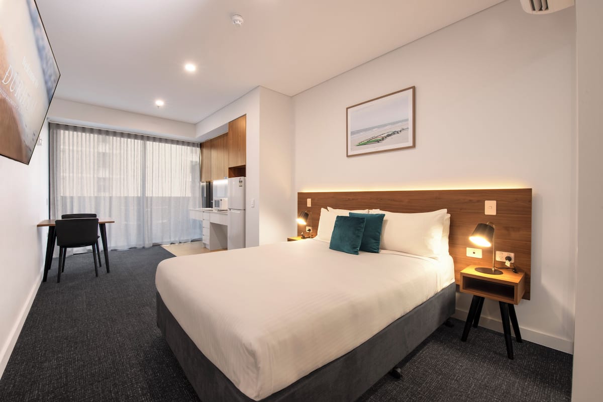 Quarto de hotel com cama, mesas de cabeceira, escrivaninha, cadeira, televisão e janela com cortinas.