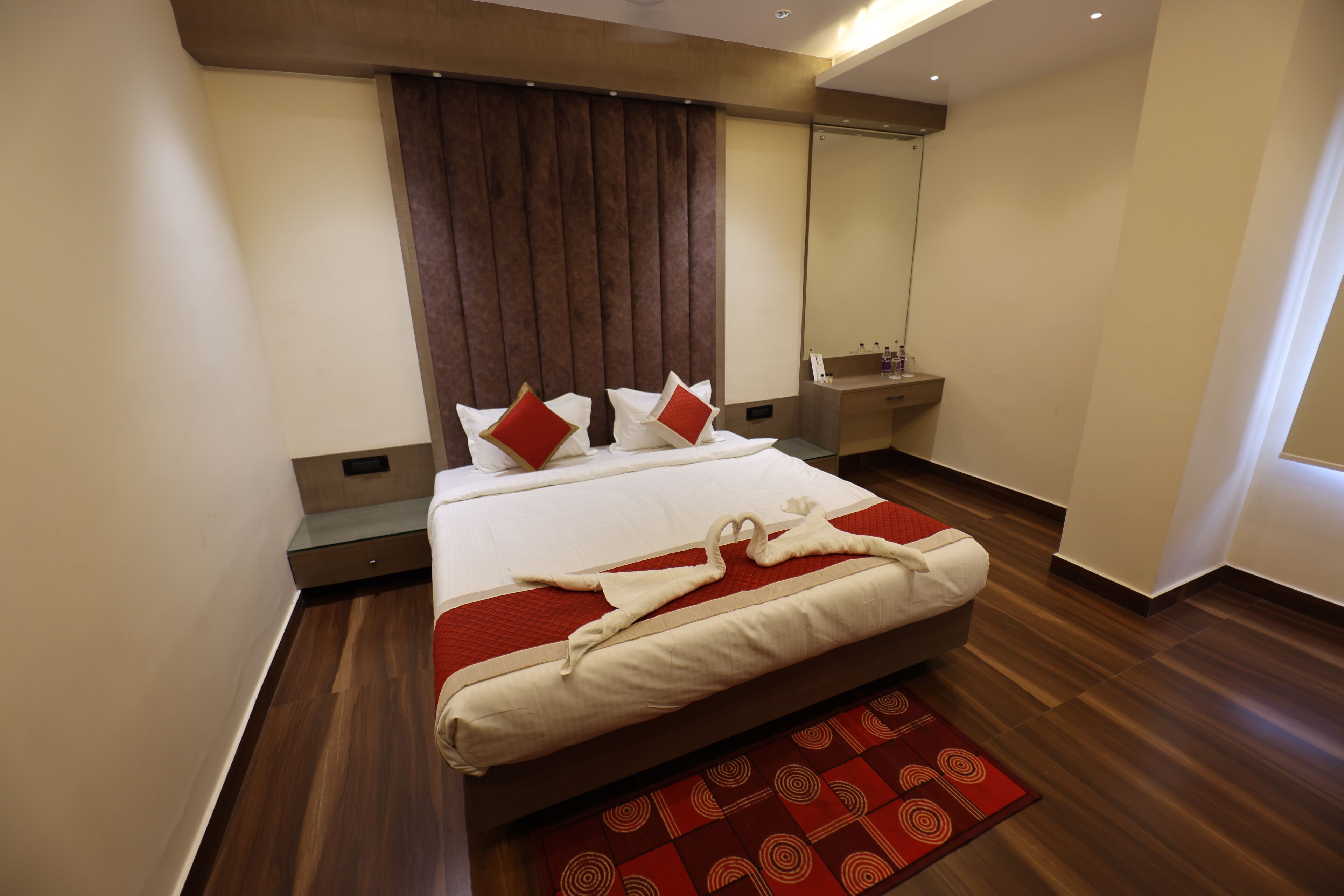Quarto de hotel moderno com uma cama grande, piso de madeira, tapete vermelho estampado e um espelho grande sobre a cama.
